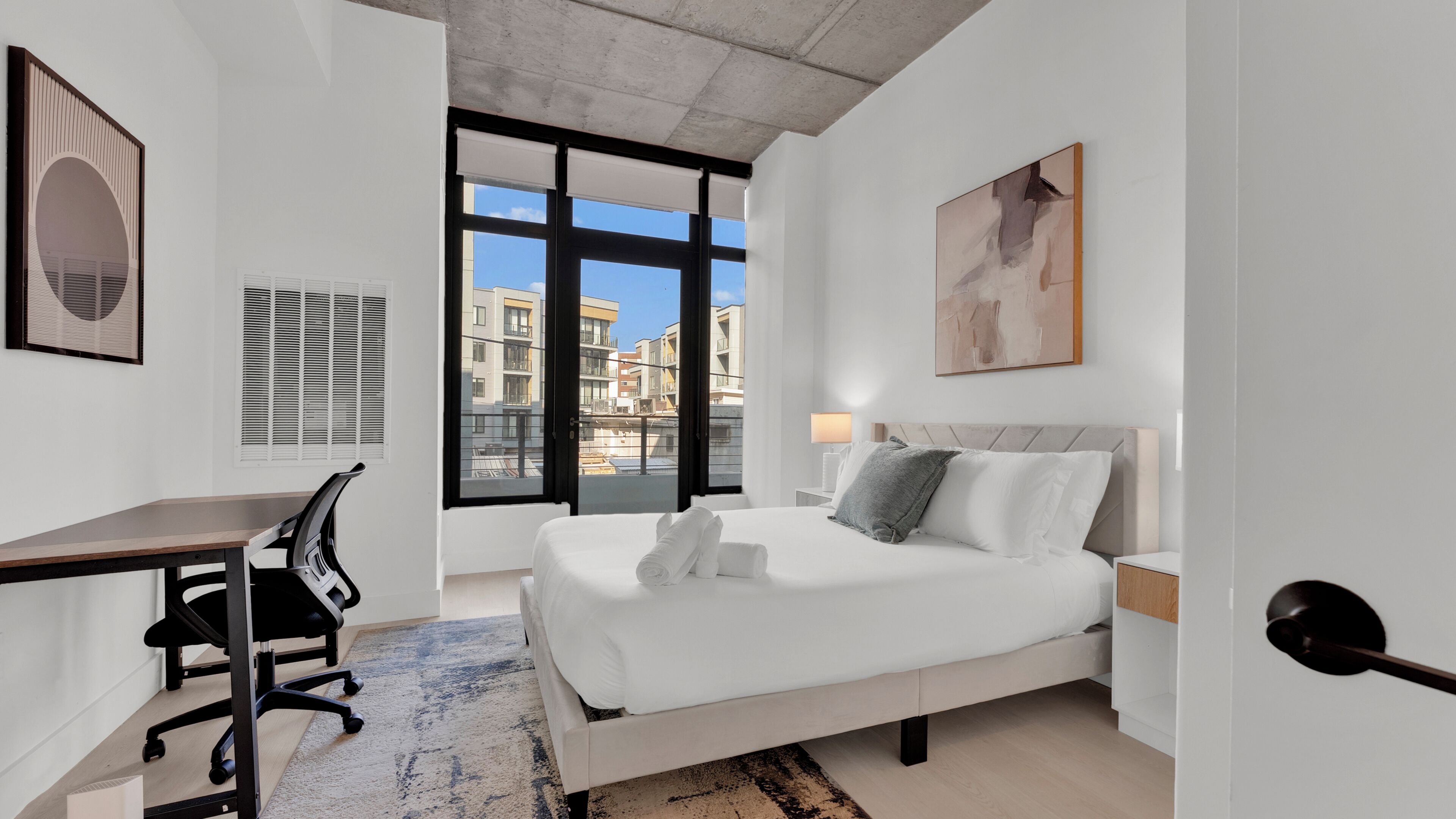 Quarto de hotel com cama, escrivaninha, cadeira e janelas amplas.