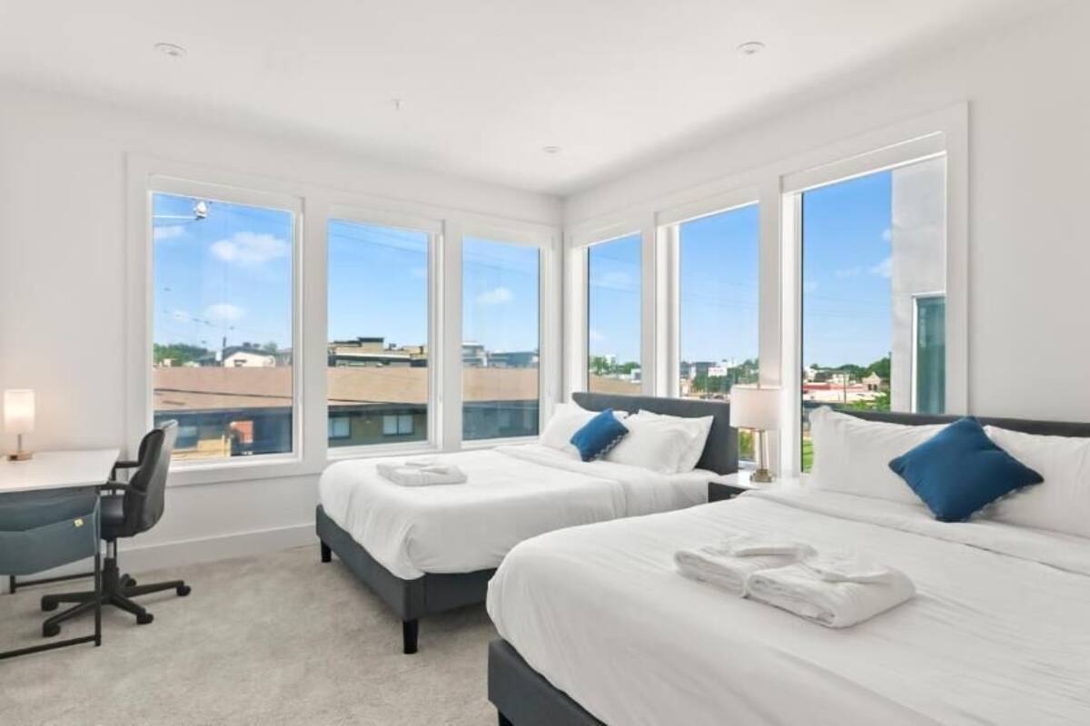 Quarto de hotel com duas camas, uma escrivaninha e grandes janelas com vista para a cidade.