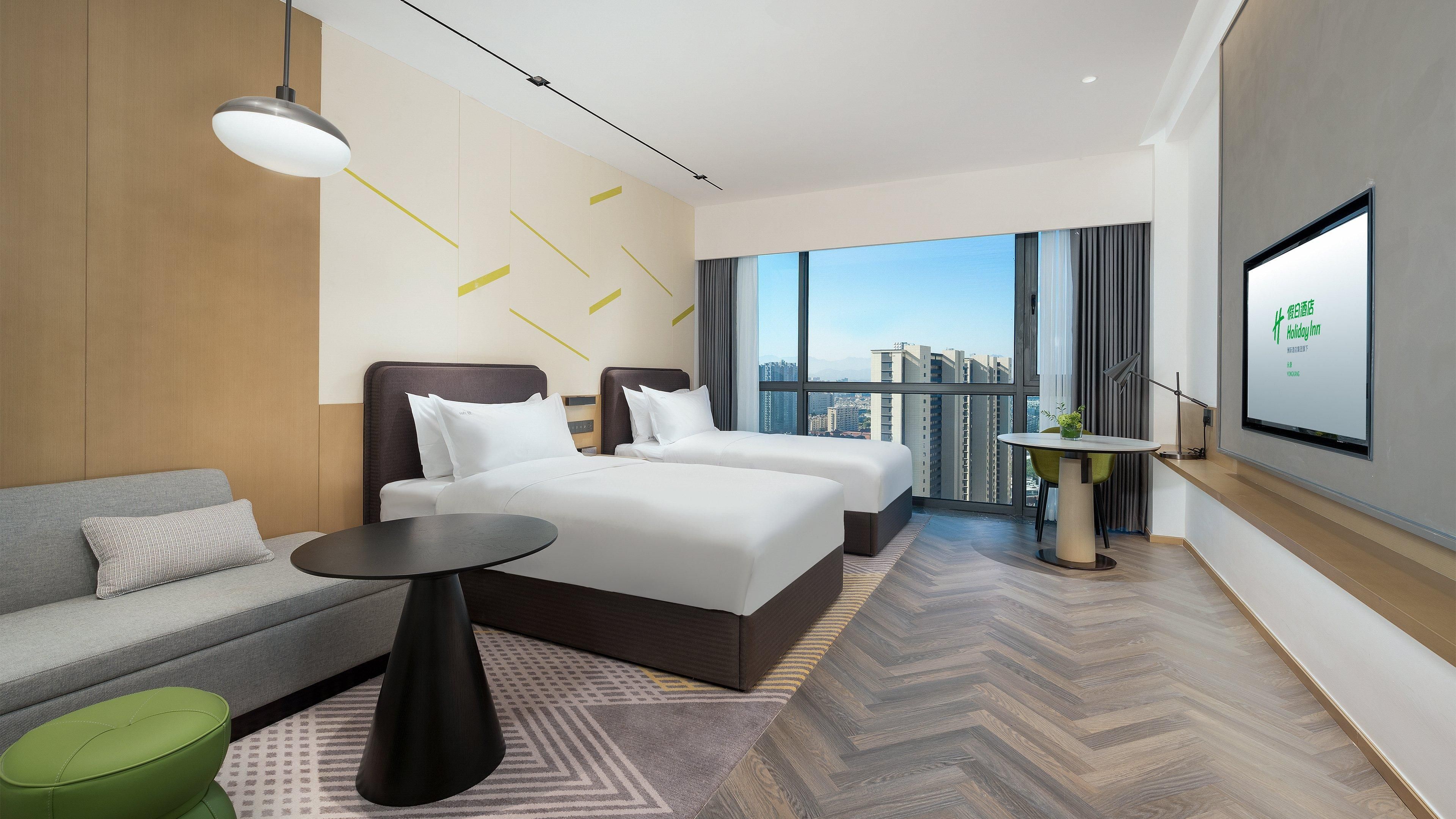Quarto de hotel moderno com uma cama grande, um sofá, uma pequena mesa e vista para a cidade através de grandes janelas.