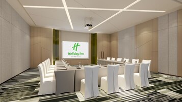 Uma sala de conferências com uma tela grande exibindo o logotipo Holiday Inn, cadeiras brancas e uma mesa comprida com garrafas de água.