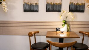 Interior de restaurante com mesa de madeira, quatro cadeiras de madeira, um vaso com flores e duas pinturas penduradas na parede.