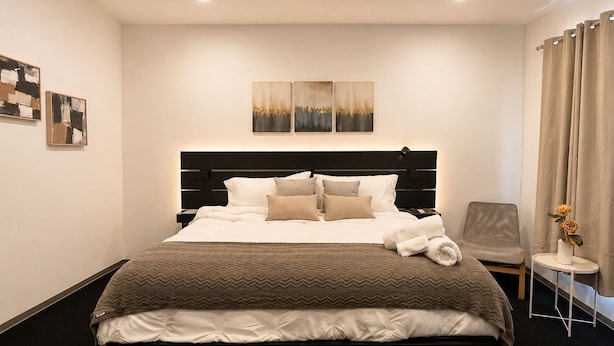 Quarto de hotel moderno com uma cama grande, cabeceira com linhas horizontais, mesinha lateral com um vaso e uma obra de arte na parede.