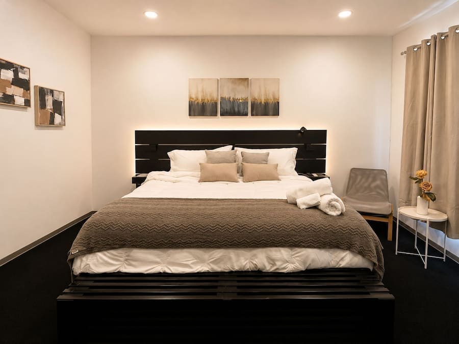 Quarto de hotel moderno com uma cama grande, cabeceira com linhas horizontais, mesinha lateral com um vaso e uma obra de arte na parede.