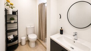 Uma casa de banho moderna com sanita branca, uma prateleira preta com produtos de higiene e um lavatório branco com espelho redondo, e cortina de chuveiro.