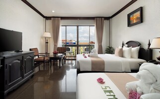 Quarto de hotel com televisão, duas camas, escrivaninha, cadeiras, abajur e vista para a cidade.