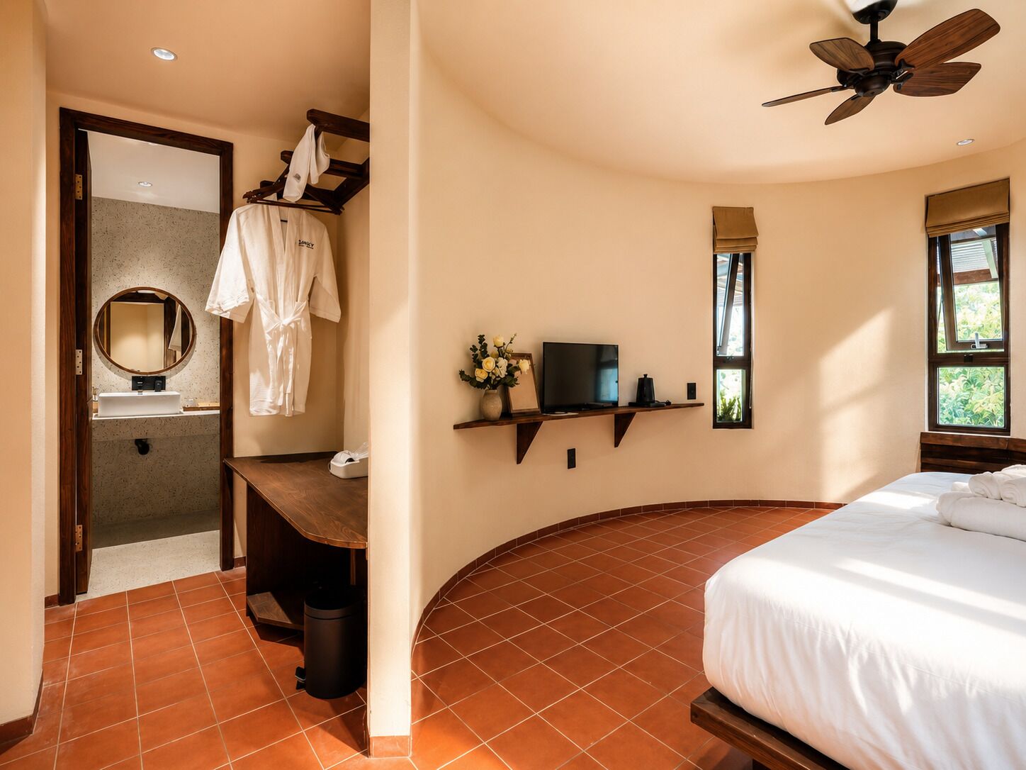 Quarto com cama, televisão, prateleira com flores, ventilador de teto e banheiro com espelho e roupão.