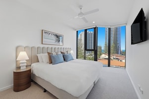 Quarto com cama, luminárias de cabeceira, ventoinha no teto, uma janela grande com vista para a cidade e TV na parede.