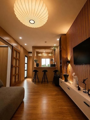 Um interior moderno com piso de madeira, um sofá, uma área de refeições com banquetas, uma cozinha com televisão e um lustre.