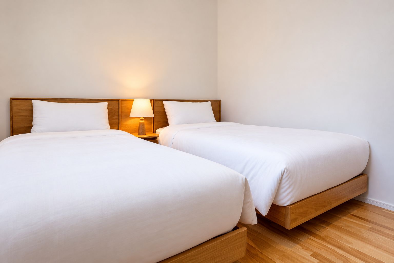 Quarto de hotel com duas camas, mesa de cabeceira com abajur e piso de madeira.