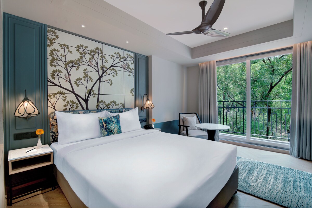 Quarto de hotel com uma cama grande, uma escrivaninha, uma cadeira, um ventilador de teto e uma janela com vista para árvores.
