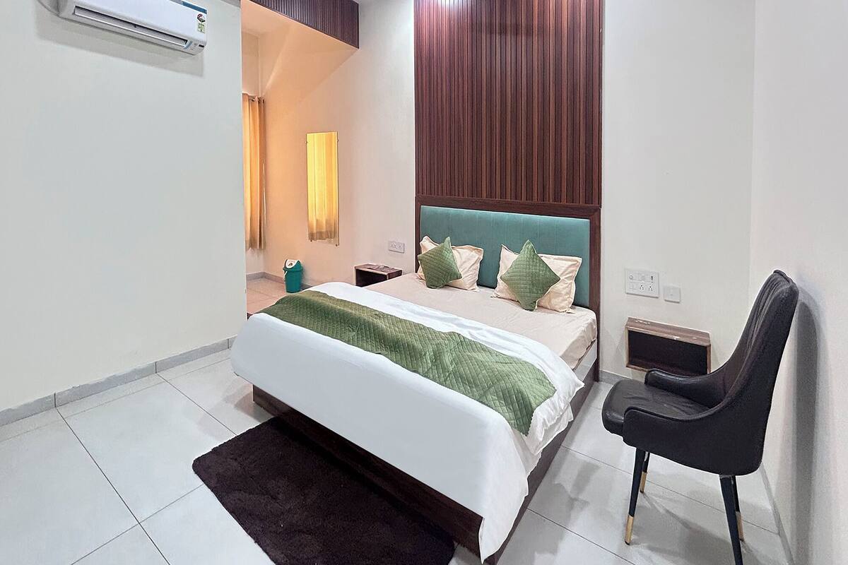 Quarto de hotel com uma cama, uma cadeira, uma mesa de cabeceira, um cesto de lixo e um ar condicionado fixado na parede.