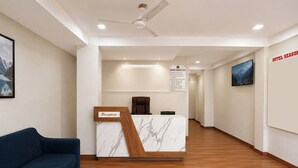 Uma área de recepção de hotel com um balcão de mármore, uma cadeira de recepcionista, um sofá azul, um ventilador de teto e uma placa de informações do hotel.