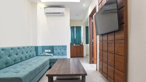 Uma sala de estar com sofá, mesa, TV fixada em um painel de madeira e um ar condicionado na parede.