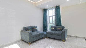 Quarto de canto com dois sofás cinzentos, uma janela com cortinas verde-água e teto branco com iluminação embutida.