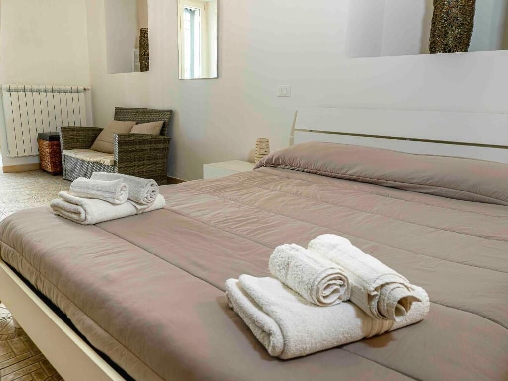 Uma cama bem arrumada com toalhas dobradas, uma cadeira de vime, uma mesinha de cabeceira e um radiador.