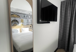 Quarto de hotel com uma cama, televisão fixada na parede e um espelho grande refletindo a fachada de um edifício.