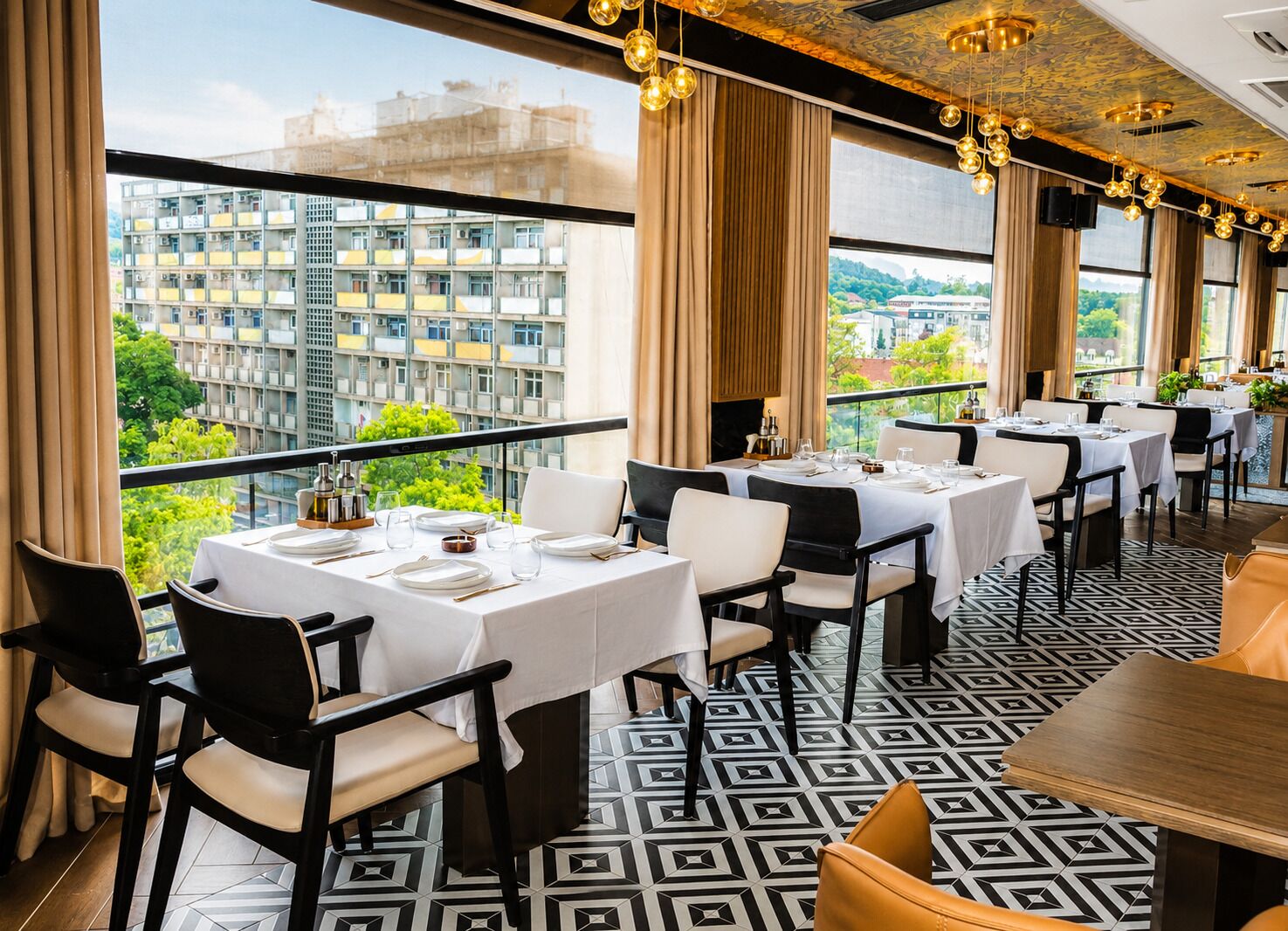 Restaurante com mesas preparadas para refeições, grandes janelas com vista para a cidade e piso estampado.