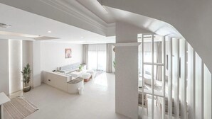 Uma sala de estar ampla com sofá, mesa de centro e vista para um quarto através de um painel de vidro.