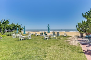 Uma área gramada com mesas e cadeiras ao ar livre e guarda-sóis, um caminho que leva à praia, e vista para o oceano.