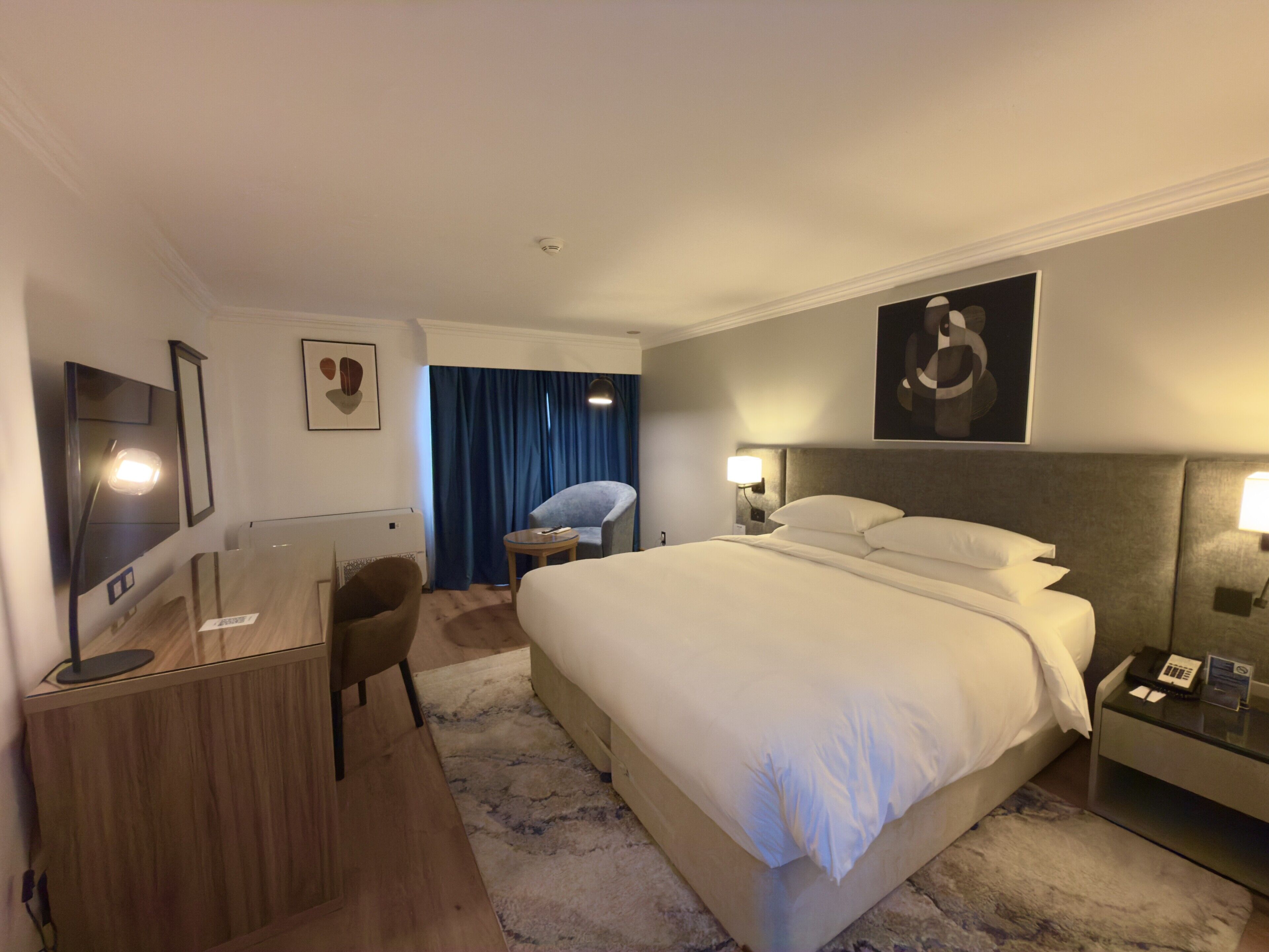 Quarto de hotel com cama, escrivaninha, cadeira, televisão e um quadro na parede.