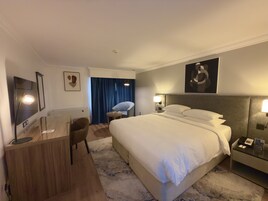 Quarto de hotel com cama, escrivaninha, cadeira, televisão e um quadro na parede.