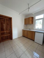 Uma cozinha com armários de madeira, bancada branca, janela e piso ladrilhado.