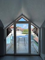 Quarto com teto triangular, varanda com mesa e cadeiras, e vista para edifícios e vegetação.