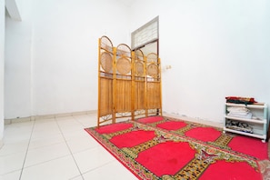 Um quarto com piso de azulejos, um tapete vermelho estampado, uma prateleira branca com roupas dobradas e um painel de madeira com desenhos circulares.