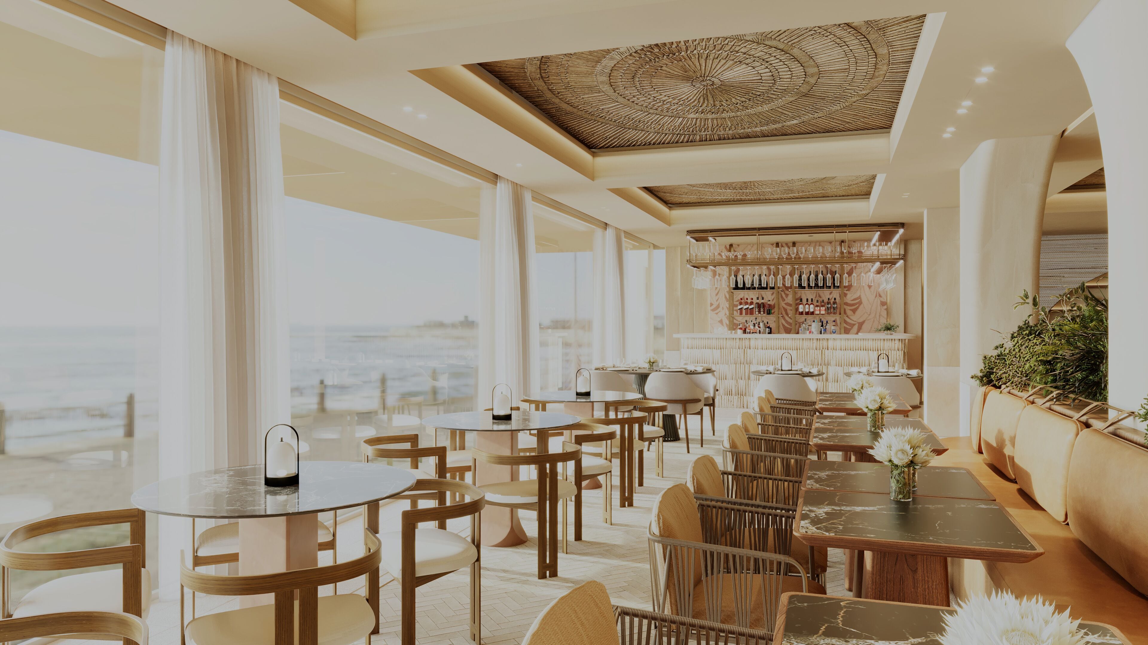 Um restaurante à beira-mar com mesas redondas, cadeiras de vime e um bar com garrafas e copos.