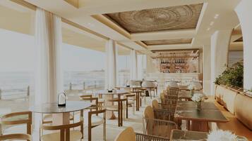 Um restaurante à beira-mar com mesas redondas, cadeiras de vime e um bar com garrafas e copos.
