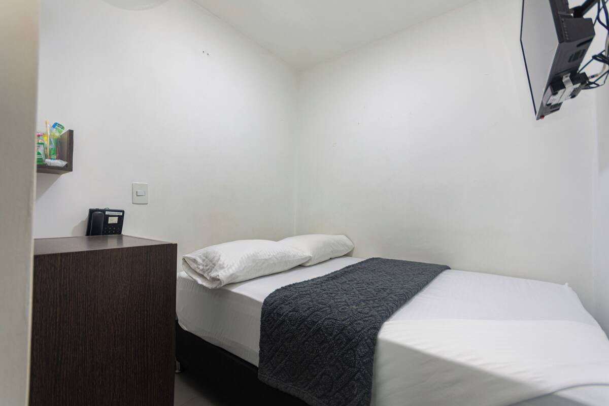 Quarto pequeno com cama de casal, escrivaninha de madeira, roupa de cama branca e manta escura.