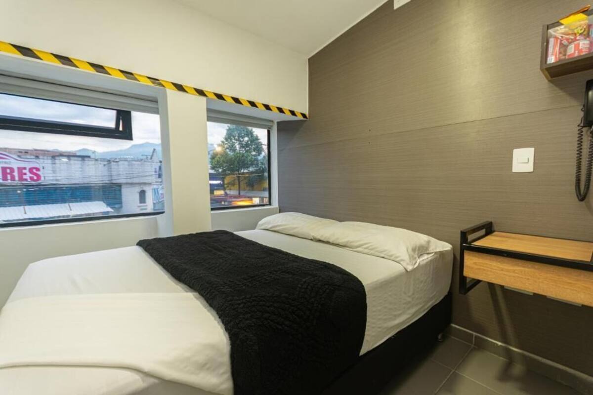 Quarto de hotel com cama, mesa de cabeceira, telefone e janela com vista para o exterior.