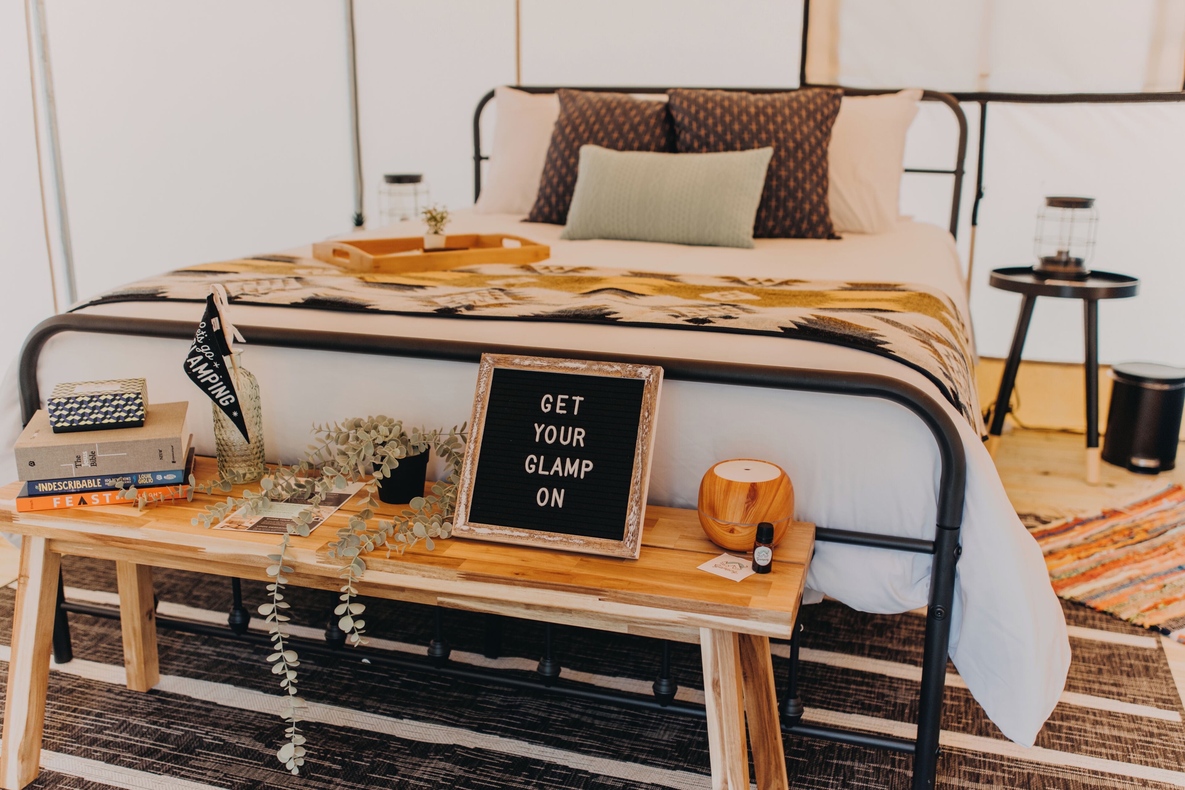Cama arrumada, placa, mesa de madeira com livros e bandeja, e uma pequena planta.