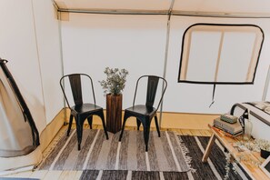 Quarto com duas cadeiras pretas, uma pequena mesa de madeira, uma planta em vaso, um tapete listrado e uma janela com moldura preta.