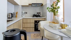 Uma cozinha compacta com cafeteira, chaleira e forno pequeno. Há uma área de refeições com uma mesa posta para dois, e um vaso de planta no parapeito da janela.