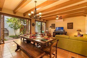 Uma área de refeições com mesa de madeira posta para uma refeição, um sofá verde e uma televisão ao fundo.