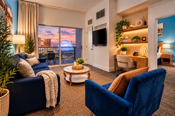 Quarto de hotel moderno com varanda, TV, escrivaninha com cadeira e área de estar com sofás e mesa de centro.