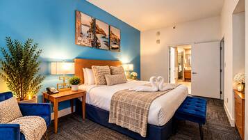 Quarto de hotel com uma cama grande, uma mesa de cabeceira com abajur, uma cadeira, um vaso de planta e uma parede com três quadros.