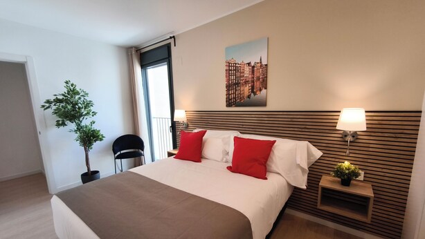 Quarto de hotel com uma cama grande, uma planta em vaso, uma cadeira, uma mesa de cabeceira com abajur e uma pintura com vista para a cidade na parede.