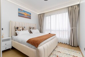 Quarto moderno com cama, mesas de cabeceira, um criado-mudo, um quadro e uma janela grande com cortinas.