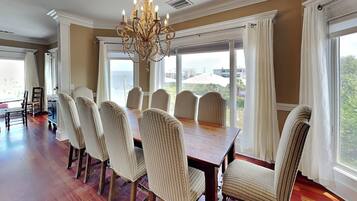 Uma sala de jantar com uma grande mesa de madeira e seis cadeiras estofadas, um candelabro e janelas amplas com cortinas.