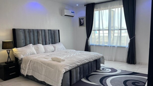 Quarto moderno com cama estofada cinza, roupa de cama branca, mesa de cabeceira preta com abajur, janela grande com cortinas escuras e tapete estampado.