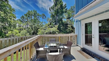 Um terraço com mesa de vidro e cadeiras de vime, cercado por uma cerca de madeira, com árvores e um edifício ao fundo.