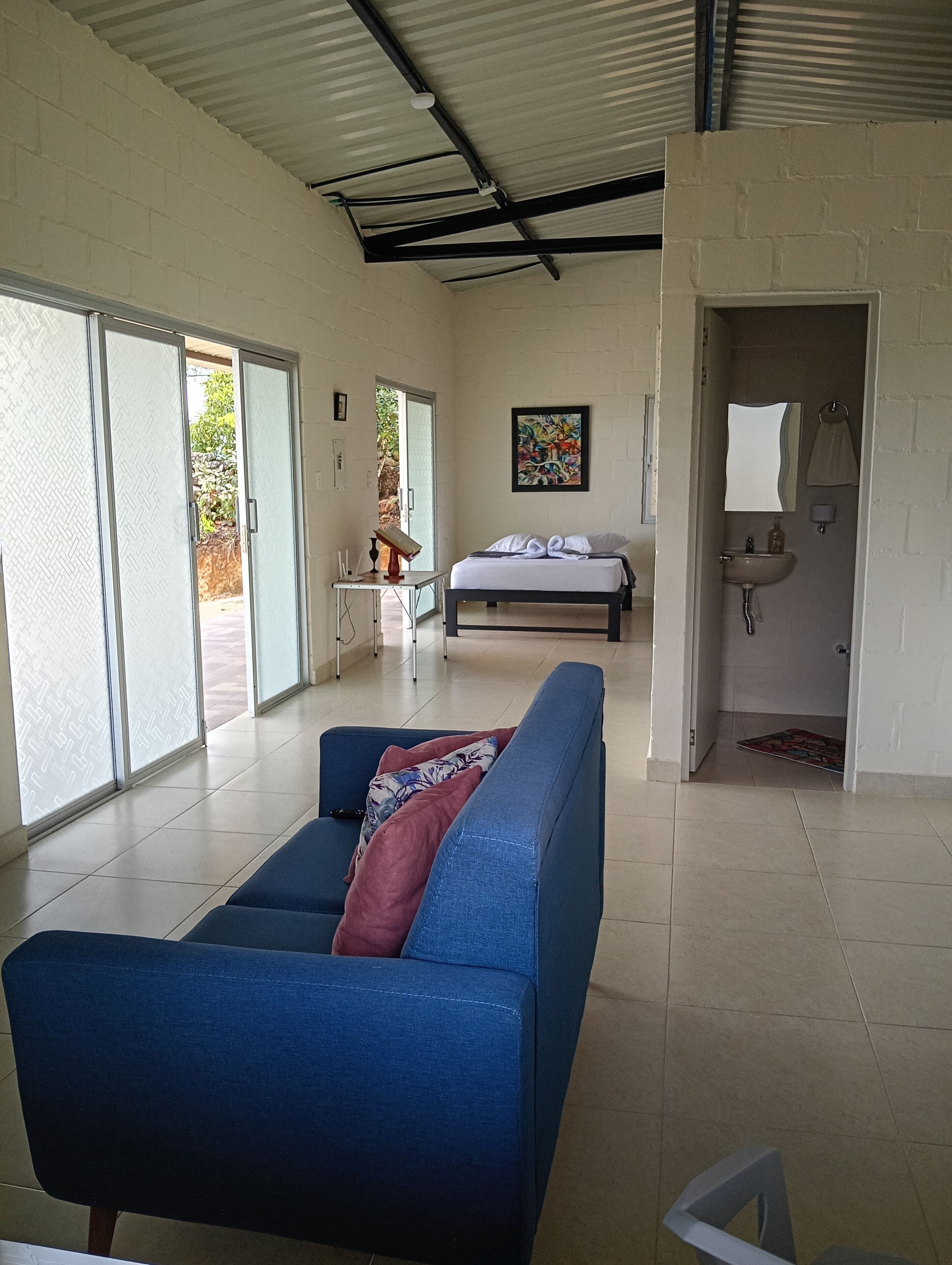 Quarto com cama, sofá azul, mesinha, quadro e banheiro com pia e espelho.