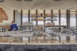Uma área de bar com um bartender, prateleiras cheias de garrafas de bebidas e vários copos.