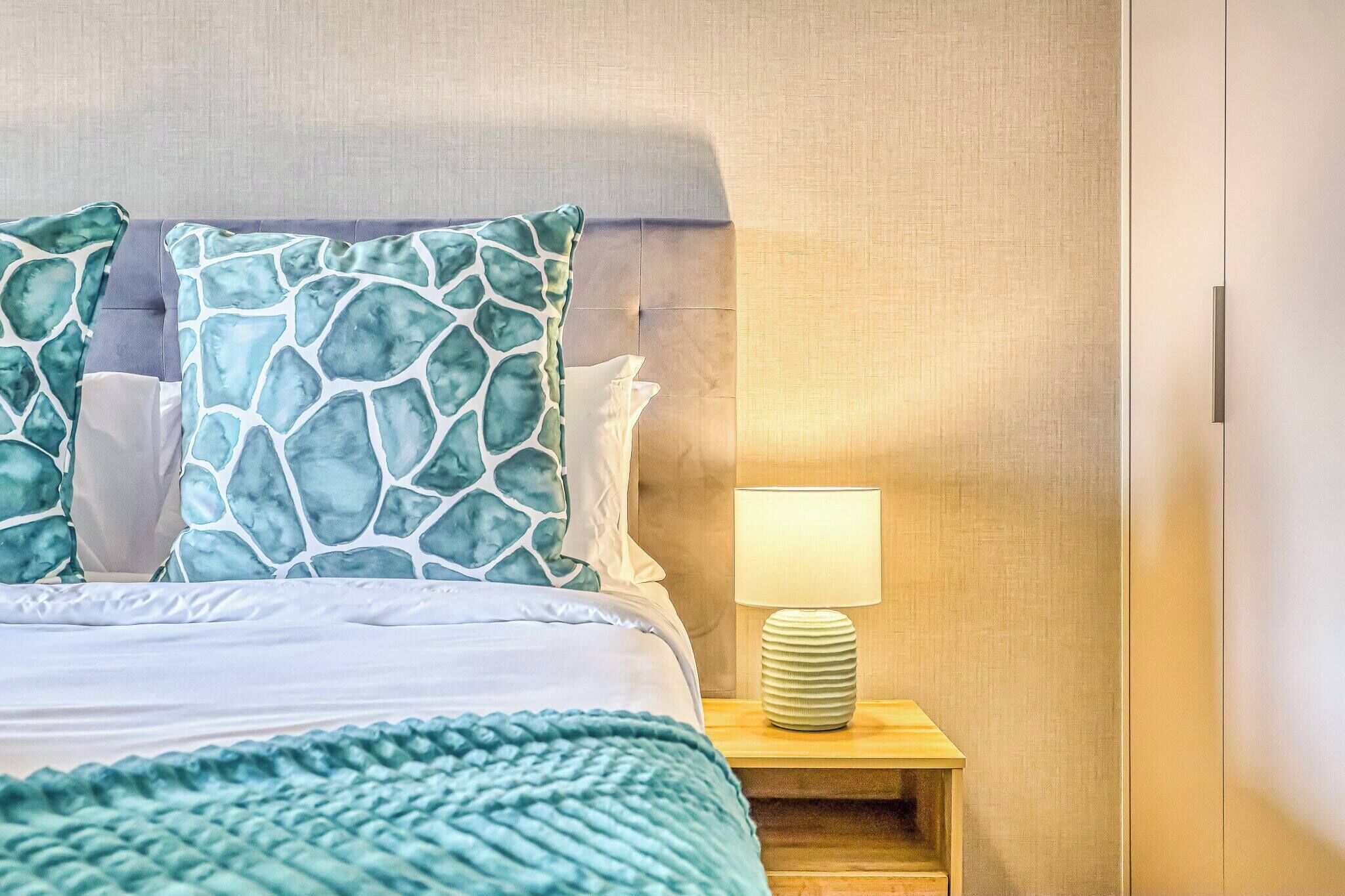 Uma cama bem arrumada com almofadas estampadas, uma luminária de cabeceira e uma mesinha de cabeceira de madeira.