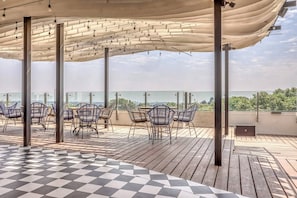 Um terraço ao ar livre com piso xadrez, cadeiras de metal, toldo e vista para a cidade.