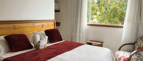 Quarto com cabeceira de madeira, cama com colcha branca e vermelha, mesa de cabeceira, cadeira e janela com cortinas.