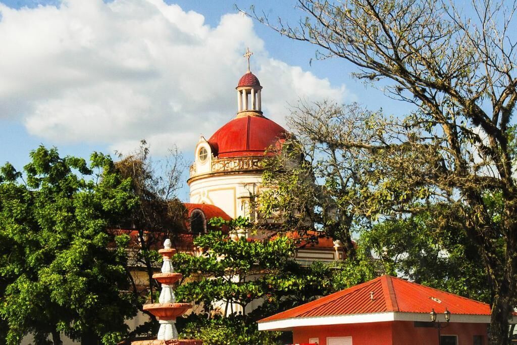 Uma igreja com cúpula vermelha e uma cruz no topo, rodeada por árvores.