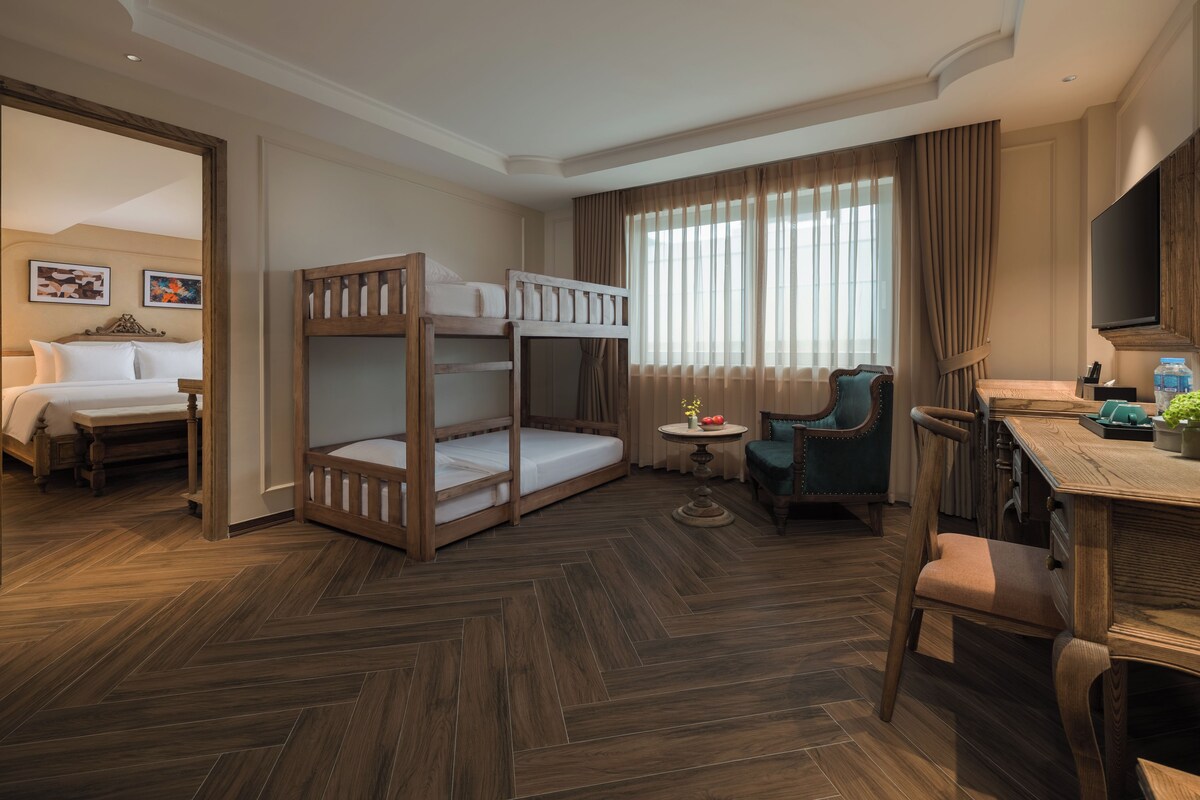 Quarto de hotel com beliche, uma escrivaninha com cadeira, televisão e uma pequena mesa com um vaso e frutas.